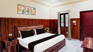 Quarto de hotel com cabeceira de madeira, cama com roupa de cama branca, mesa de cabeceira e mesa lateral com uma planta, e porta com janela.