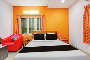 Quarto de hotel com cama, poltrona vermelha, mesa de cabeceira com abajur e janela com cortinas.