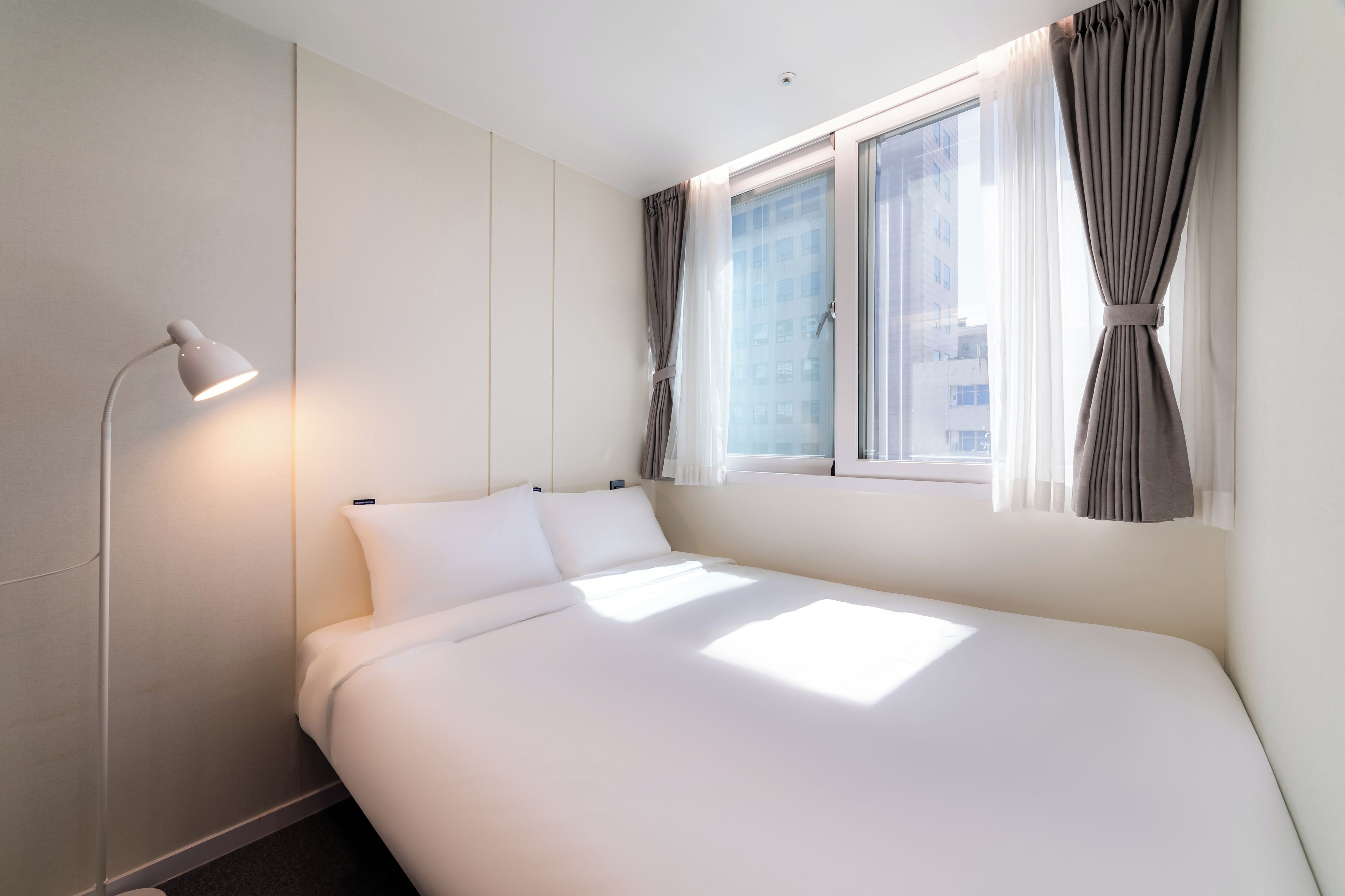 Quarto de hotel com cama, luminária de mesa e janela com cortinas.