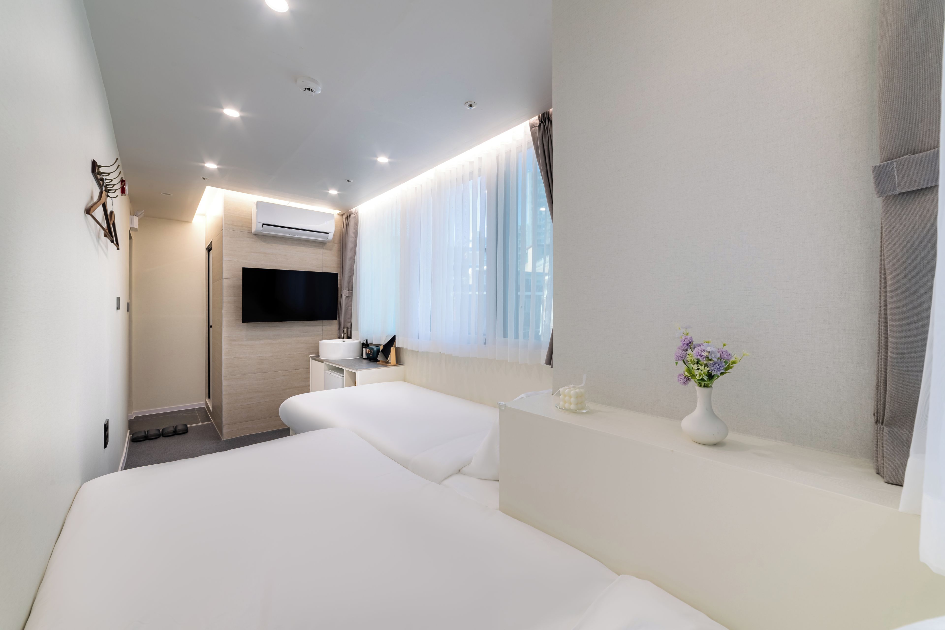 Quarto de hotel moderno com uma cama grande, televisão de tela plana na parede, mesa de cabeceira pequena e uma janela com cortinas translúcidas.