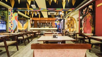 Interior de restaurante com mesas e bancos de madeira, área de bar e decorações coloridas nas paredes.