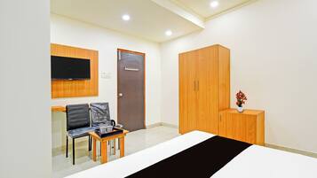 Quarto de hotel com cama, escrivaninha, cadeira, guarda-roupa e televisão na parede.