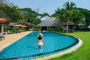Piscina com borda curva, rodeada por espreguiçadeiras e guarda-sóis, com palmeiras e um edifício ao fundo.
