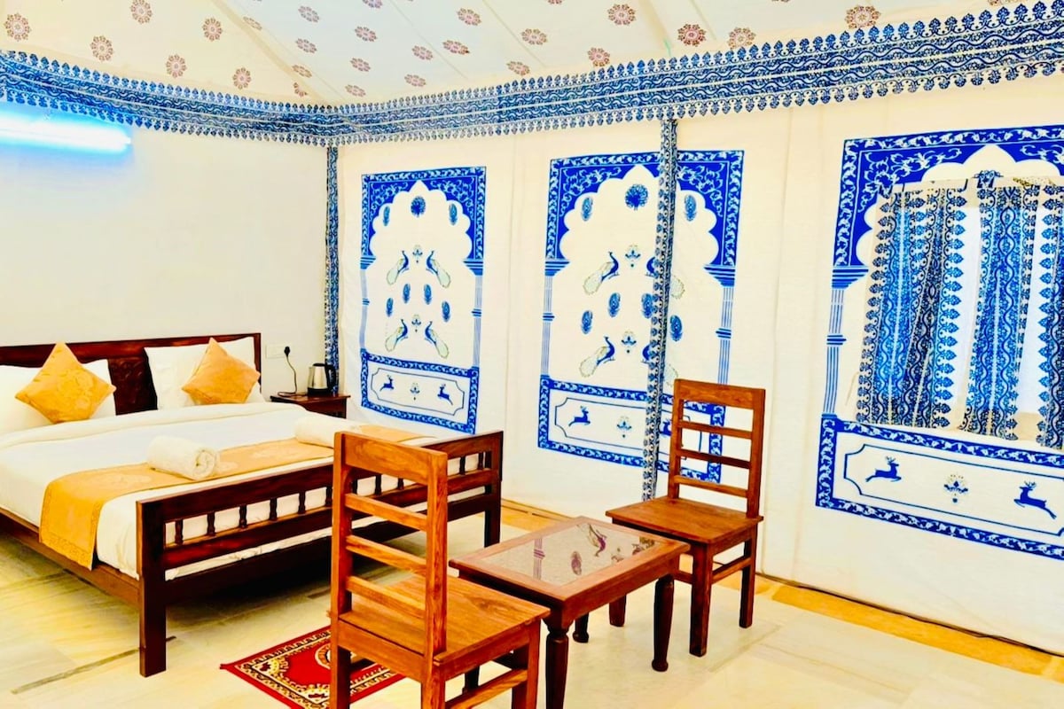 Quarto de hotel com cama, mesinhas de cabeceira de madeira, uma mesa, cadeiras e cortinas decorativas com padrão azul e branco.