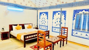 Quarto de hotel com cama, mesinhas de cabeceira de madeira, uma mesa, cadeiras e cortinas decorativas com padrão azul e branco.