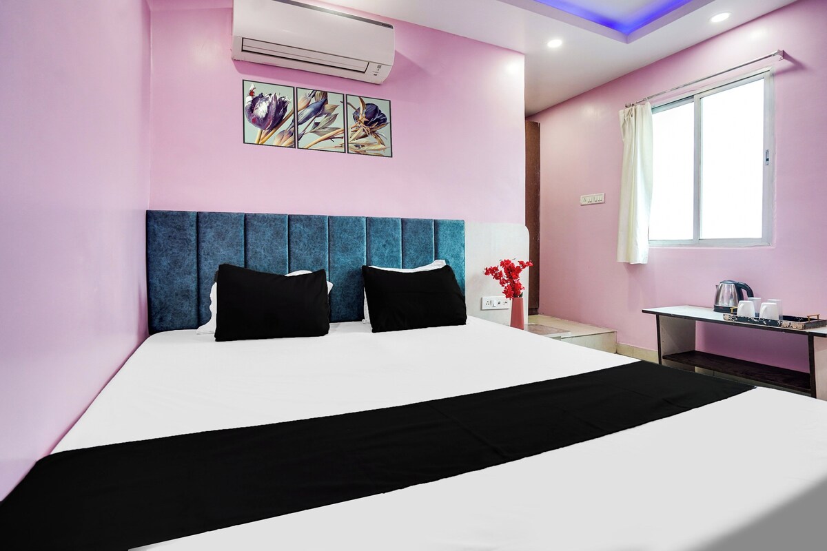 Quarto de hotel moderno com uma cama grande, cabeceira azul, pequena mesa com chaleira e janela com cortinas.