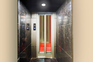 Um elevador com carpete vermelho, paredes metálicas e uma luminária no teto.