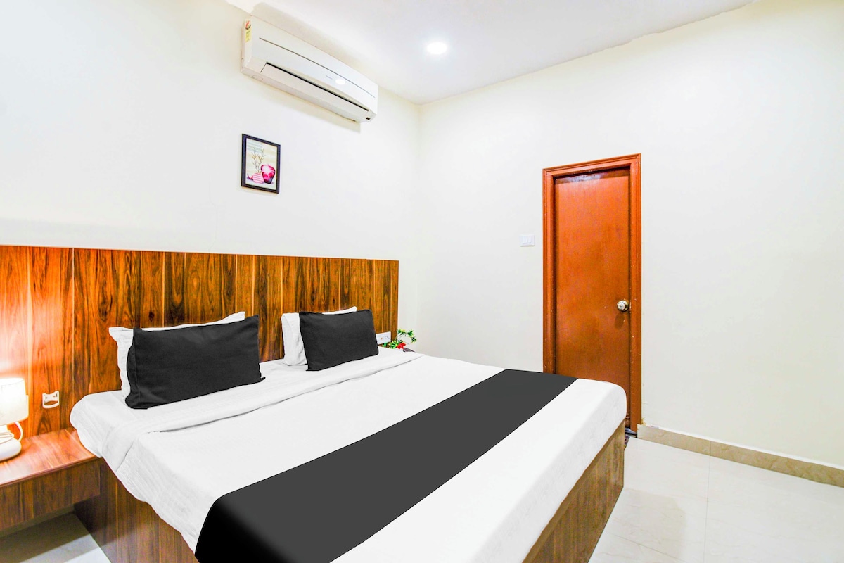 Quarto de hotel com cabeceira de madeira, cama com roupa de cama preta e branca, aparelho de ar condicionado, quadro emoldurado na parede e porta de madeira.