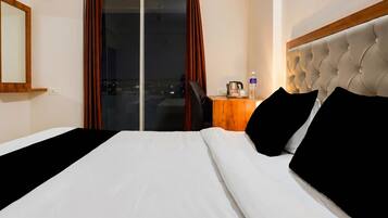 Quarto de hotel com uma cama grande, mesa de cabeceira com abajur, janela com cortinas e vista da cidade à noite.