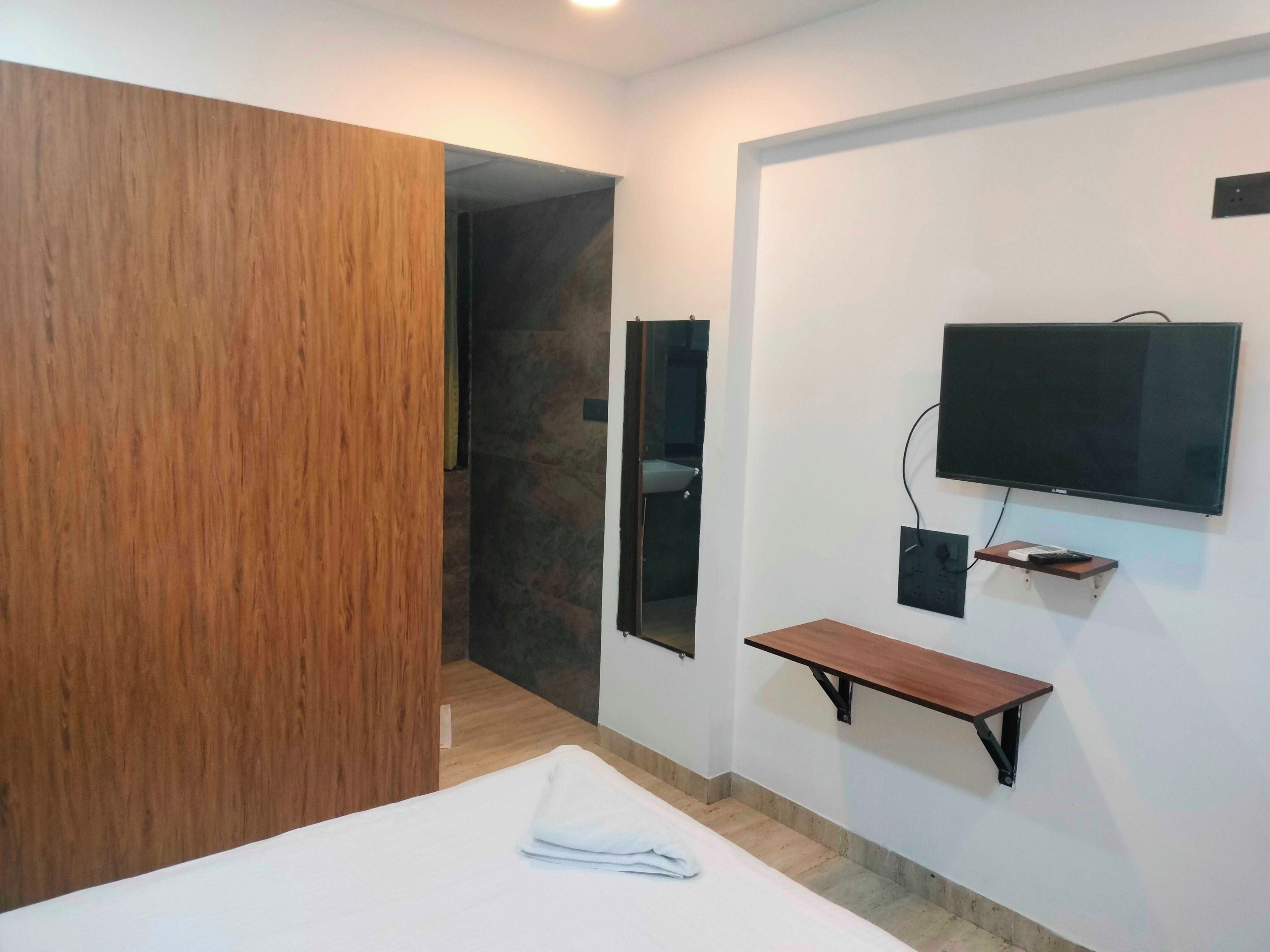 Quarto de hotel com cama, guarda-roupa de madeira, TV fixada na parede e uma pequena prateleira.