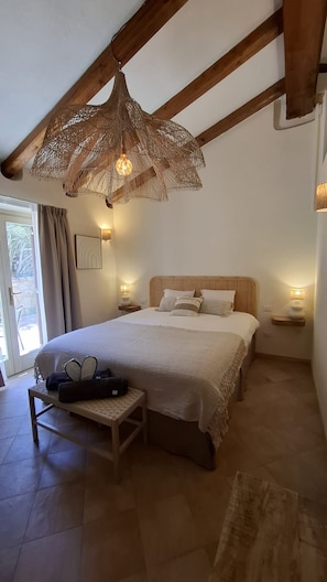 Quarto com cama grande, vigas de madeira, luminária pendente, abajures e um banco com sapatos.