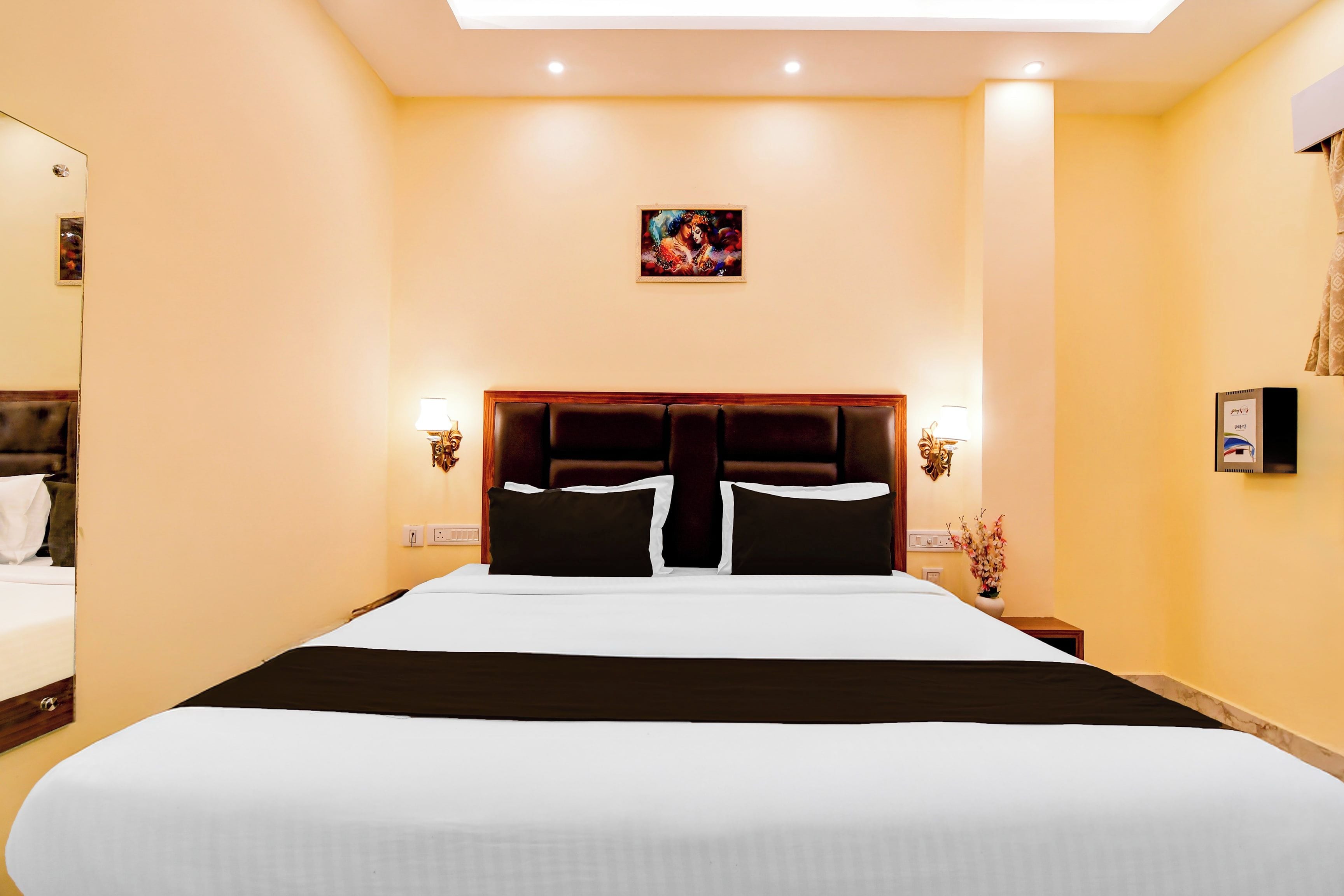 Quarto de hotel com uma cama grande, cabeceira, abajures, um quadro na parede e uma mesa de cabeceira com um vaso de flores.