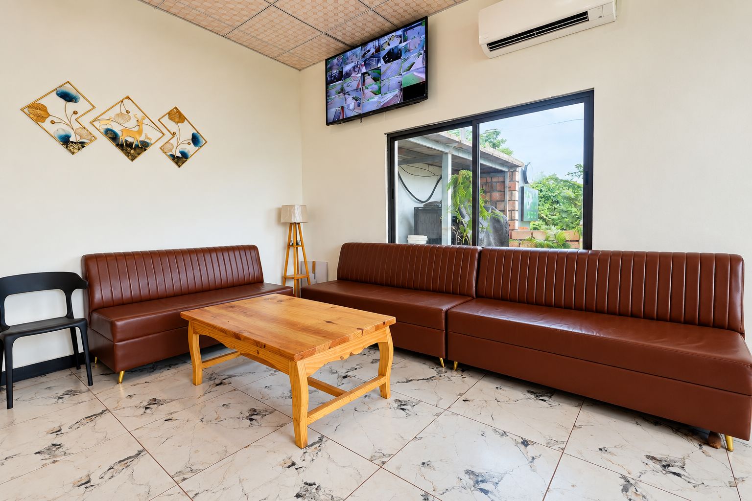 Sala de estar moderna com sofá de couro marrom, mesa de centro de madeira e uma cadeira preta. Uma televisão está instalada na parede, e há uma janela com vista para o verde lá fora.