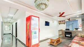 Um saguão de hotel com mesa de recepção, geladeira abastecida com bebidas, relógio e ventilador de teto.