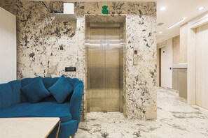 Um elevador moderno com acabamento metálico, um sofá azul com almofadas e uma parede com padrão marmorizado.