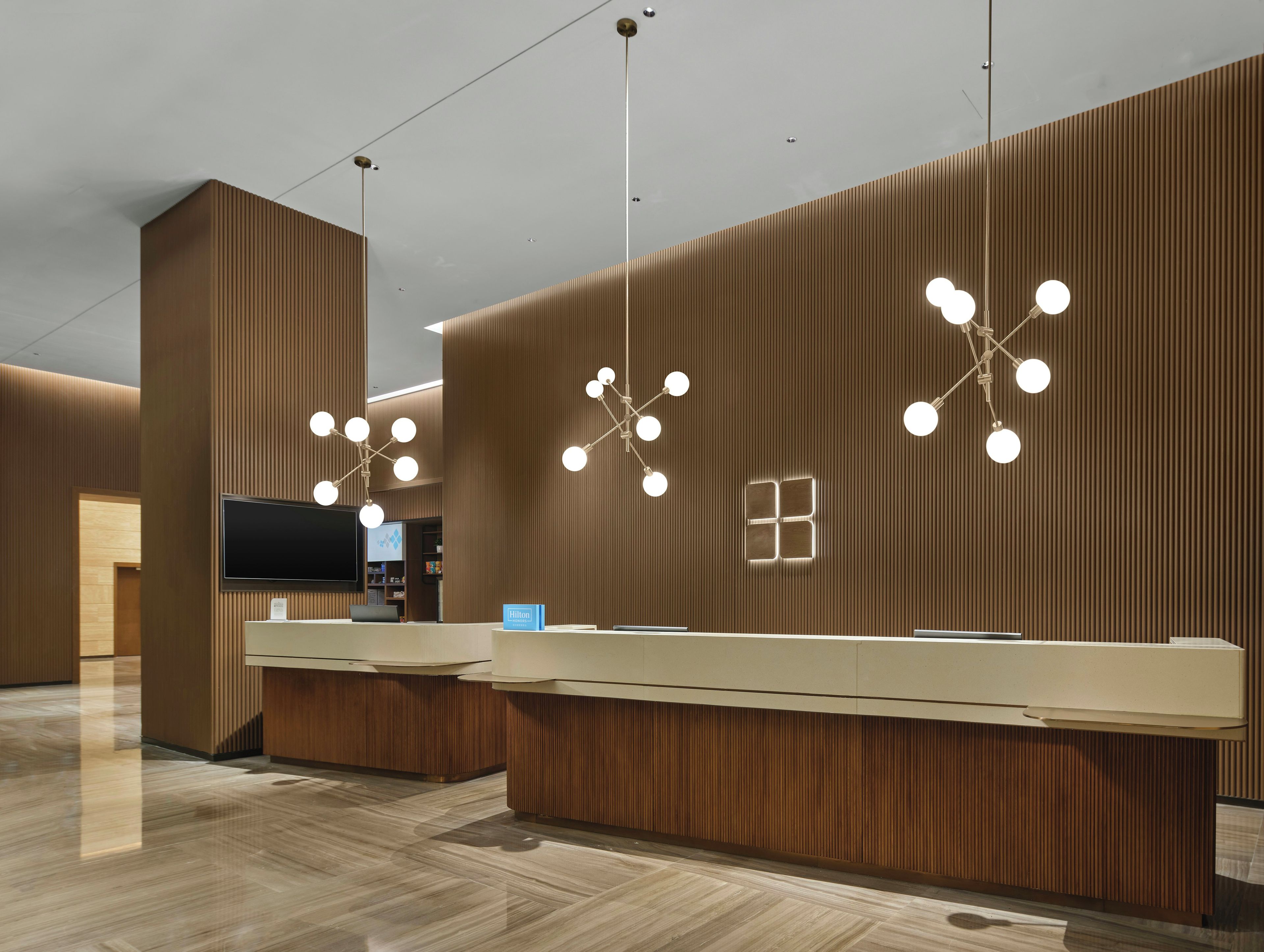 Uma área de recepção moderna em hotel, com balcão de madeira, televisão de tela plana e iluminação pendente.