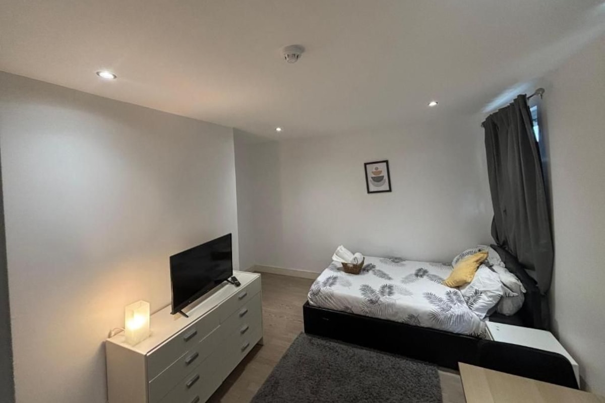 Quarto com cama, televisão sobre suporte, mesa de cabeceira e quadro pendurado na parede.