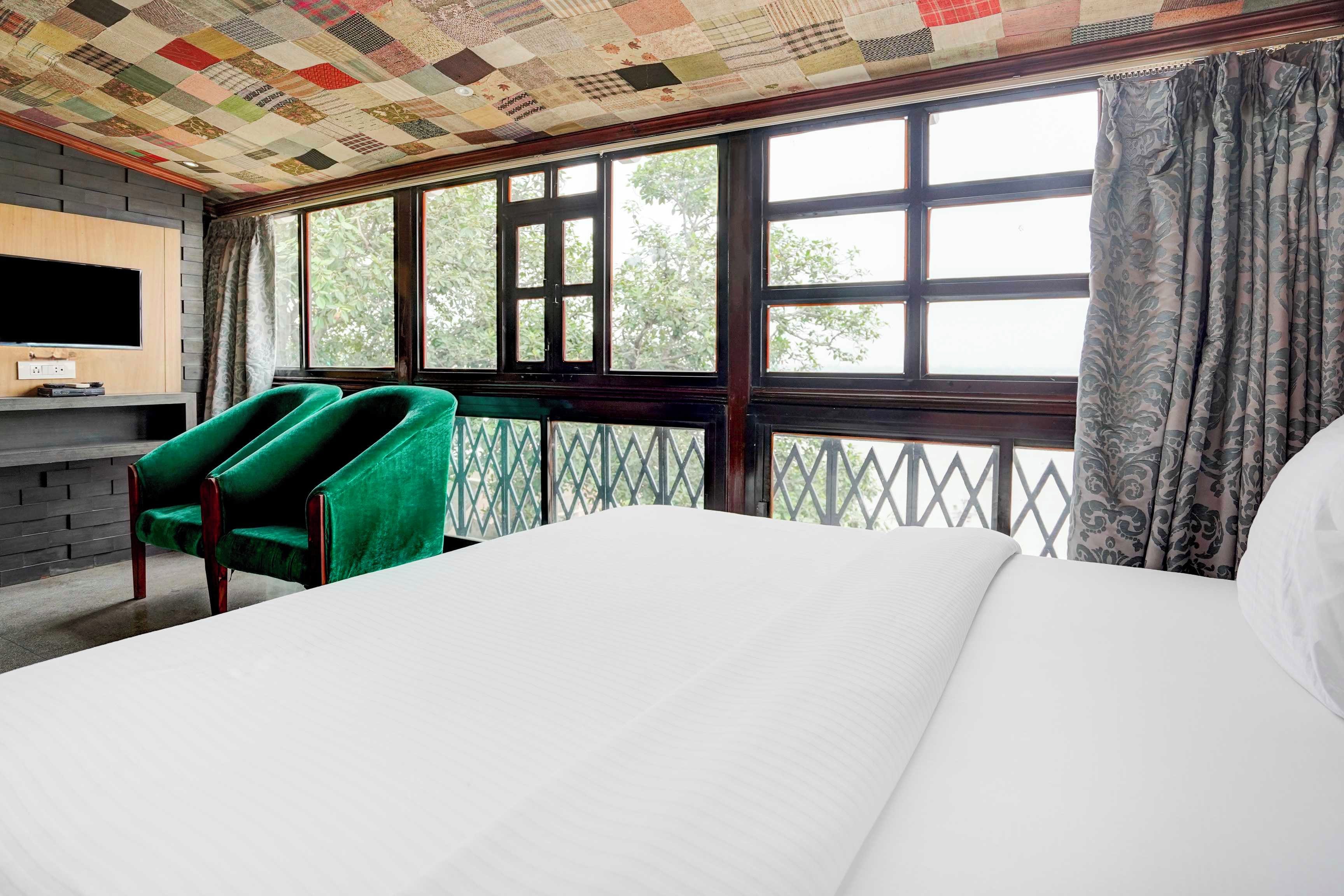 Quarto de hotel com uma cama grande, uma poltrona verde, televisão de tela plana e vista para árvores pelas janelas.