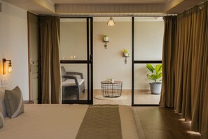 Quarto de hotel com cama, uma pequena mesa, cadeira, planta em vaso e porta de vidro corrediça para área externa.