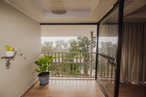 Uma varanda com piso de madeira, um vaso com planta e vista para árvores e edifícios.
