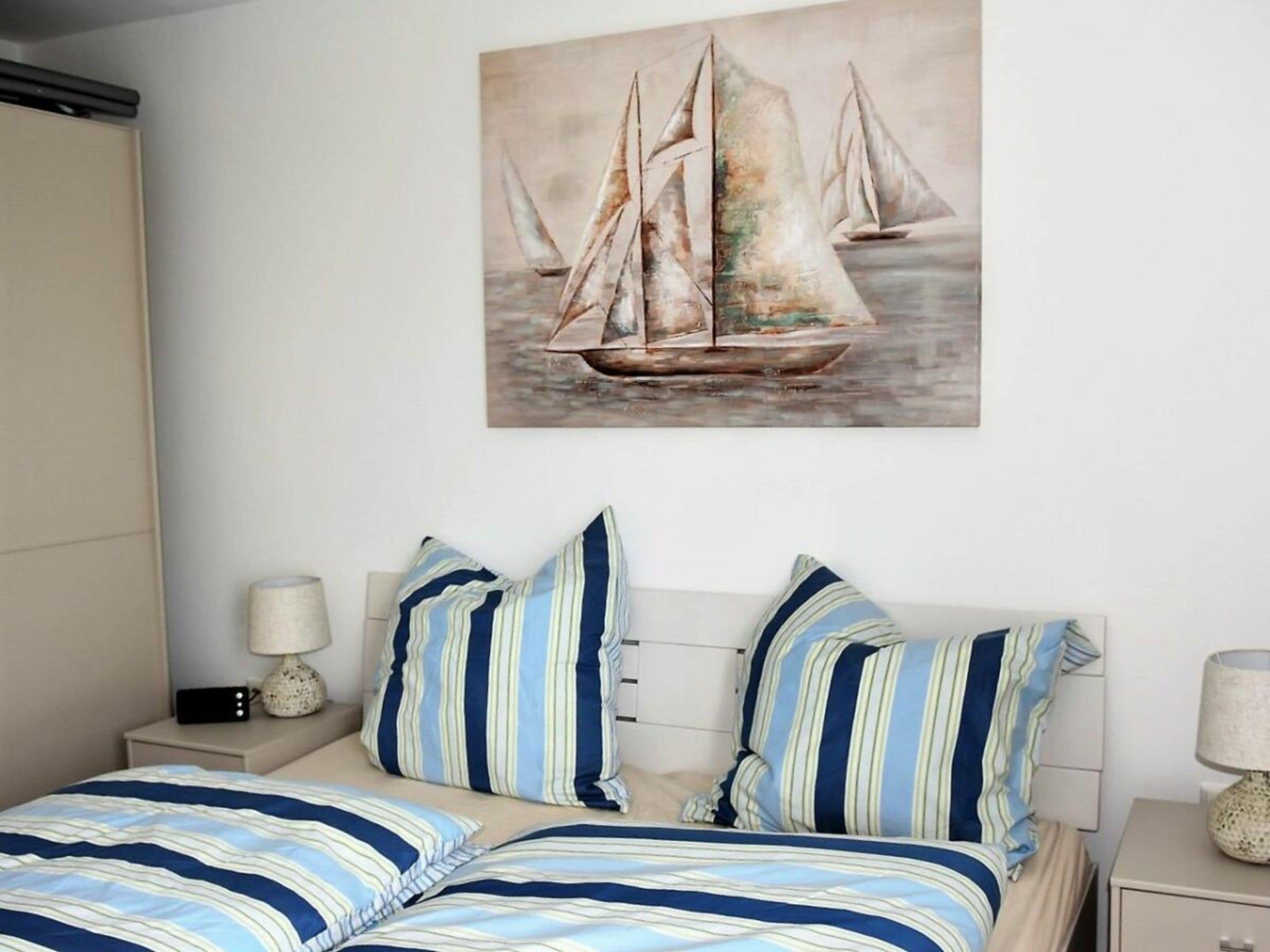 Quarto com cama, almofadas listradas, mesa de cabeceira com abajur e pintura de barcos a vela na parede.