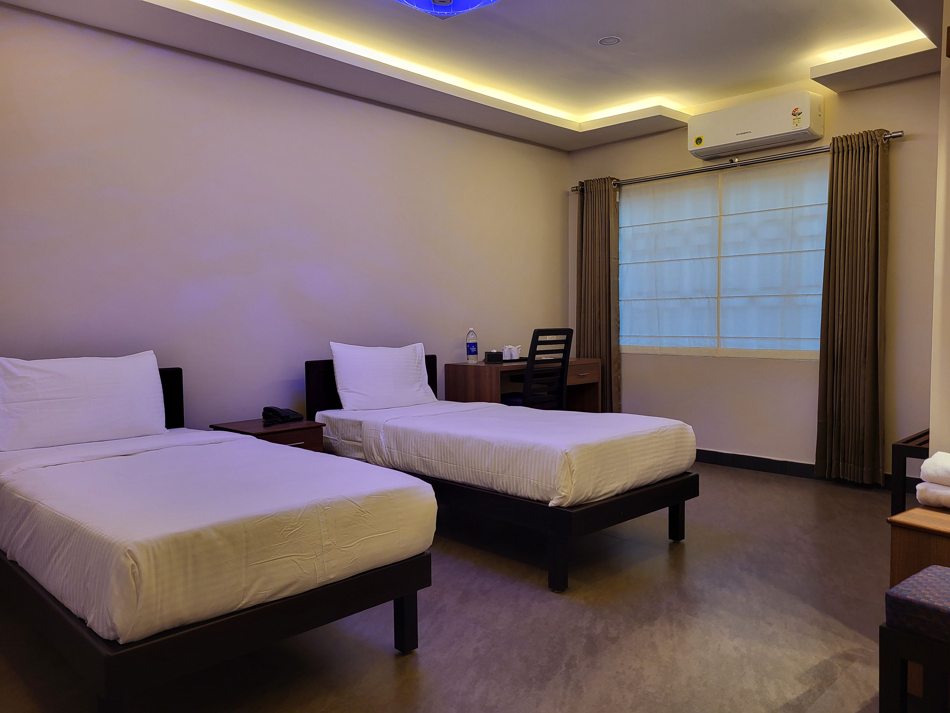 Quarto de hotel com duas camas, uma escrivaninha, uma cadeira, ar condicionado e uma janela com cortinas.