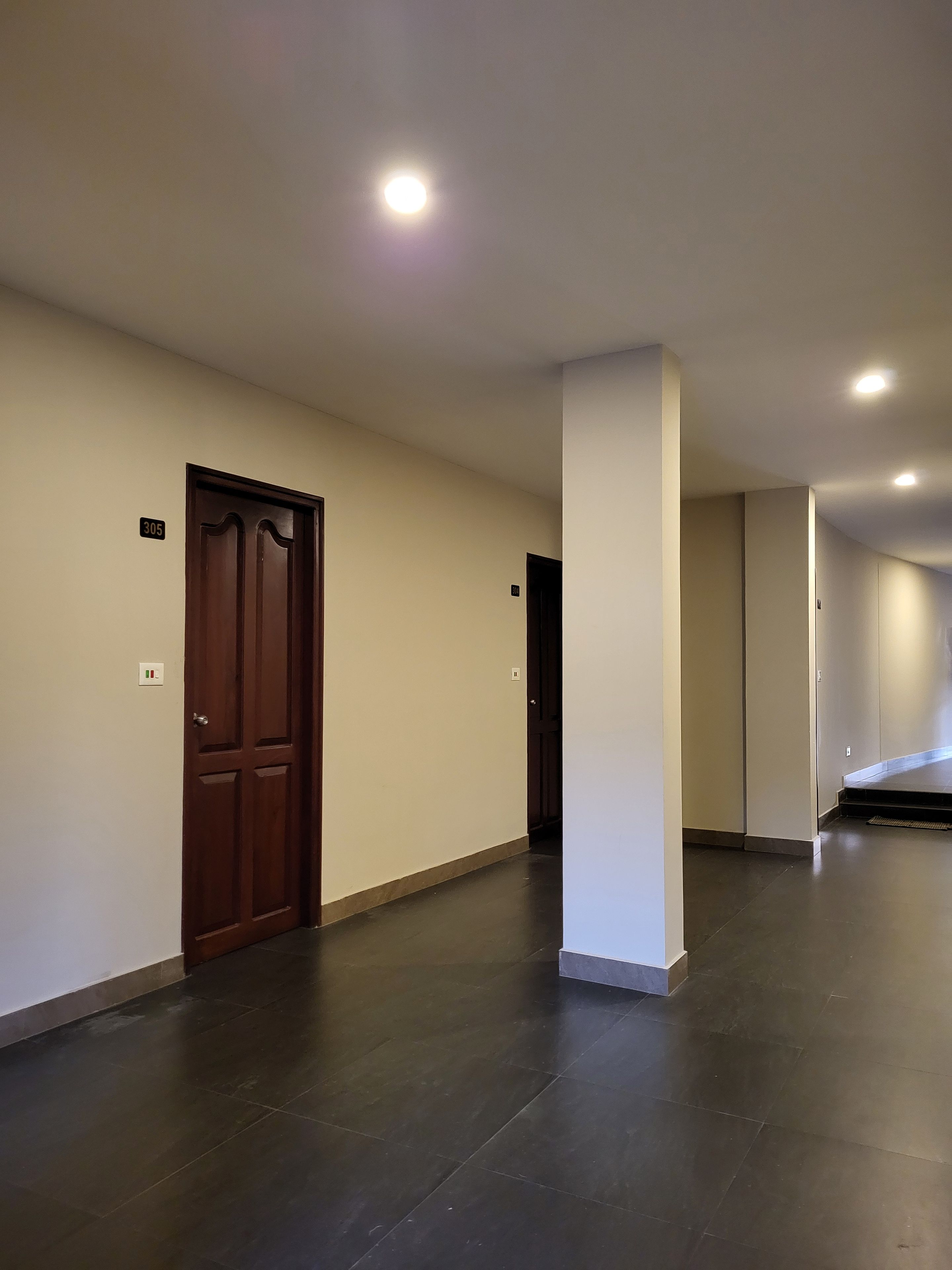 Um corredor com porta de madeira, luminárias embutidas no teto e piso ladrilhado.
