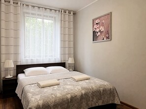 Quarto de hotel com cama, duas luminárias de cabeceira, janela com cortinas e um quadro na parede.