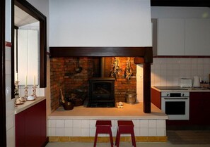 Uma cozinha com lareira de tijolos, dois banquinhos vermelhos, forno branco e painel de azulejos.