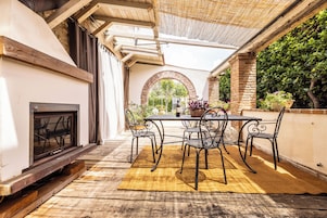 Um pátio externo com mesa e cadeiras, uma lareira e vista para um jardim através de uma porta em arco.