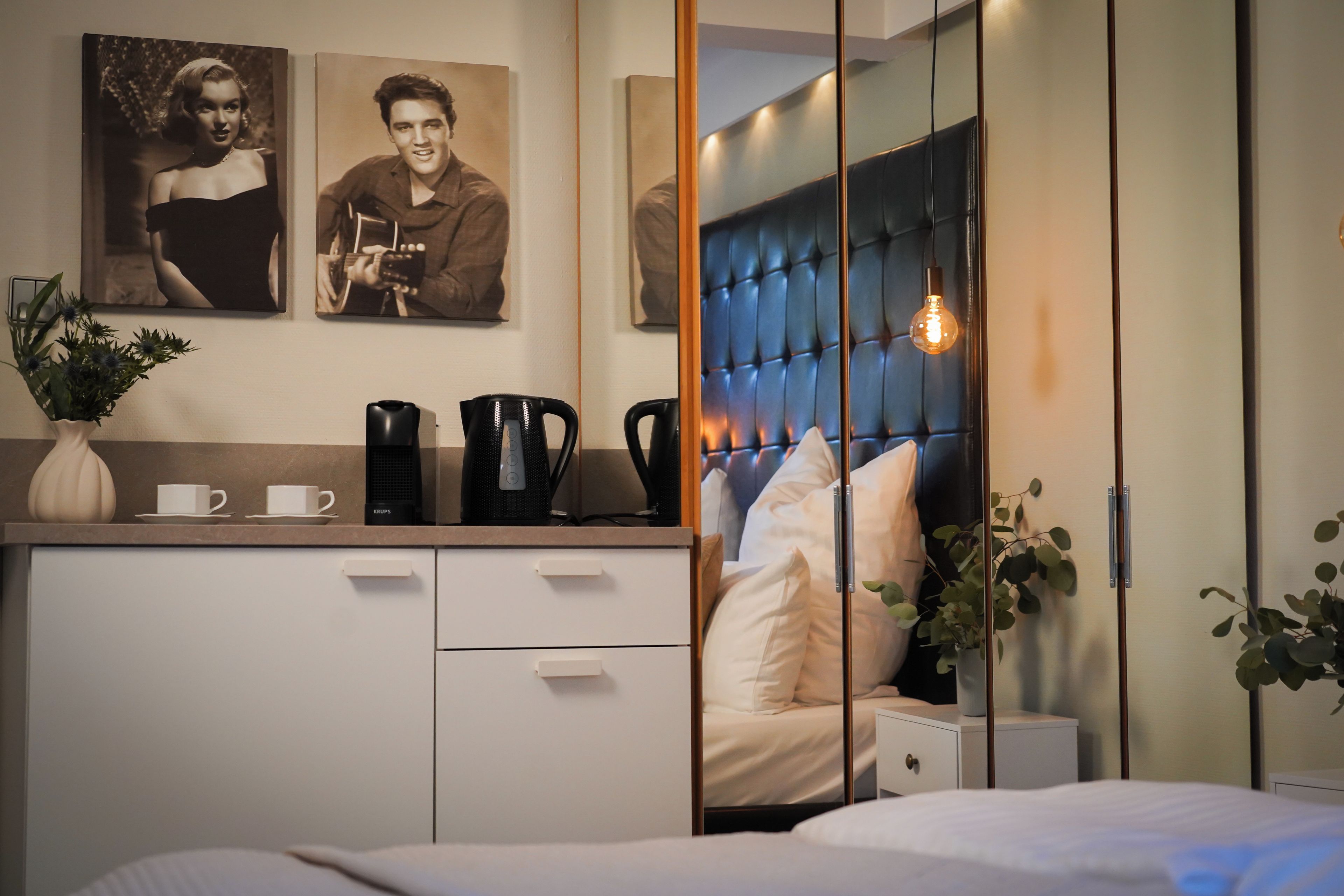 Quarto moderno com cama, mesa de cabeceira e espelho refletindo o ambiente. Quadros emoldurados na parede e um vaso com flores no penteadeira.
