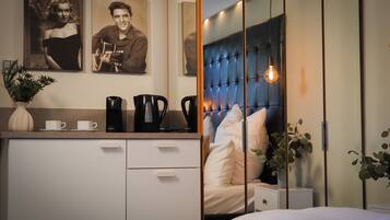 Quarto moderno com cama, mesa de cabeceira e espelho refletindo o ambiente. Quadros emoldurados na parede e um vaso com flores no penteadeira.