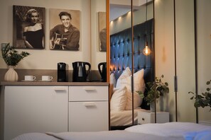 Quarto moderno com cama, mesa de cabeceira e espelho refletindo o ambiente. Quadros emoldurados na parede e um vaso com flores no penteadeira.