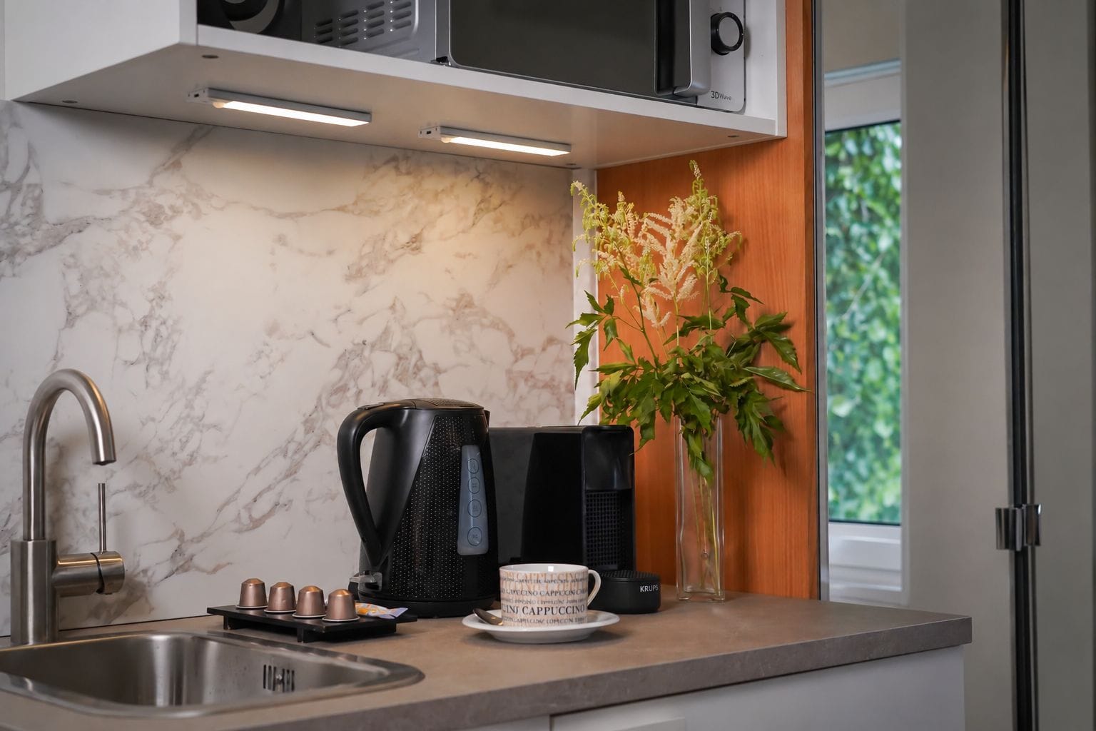 Cozinha moderna com bancada de mármore, chaleira, cafeteira, xícara e vaso com plantas verdes.