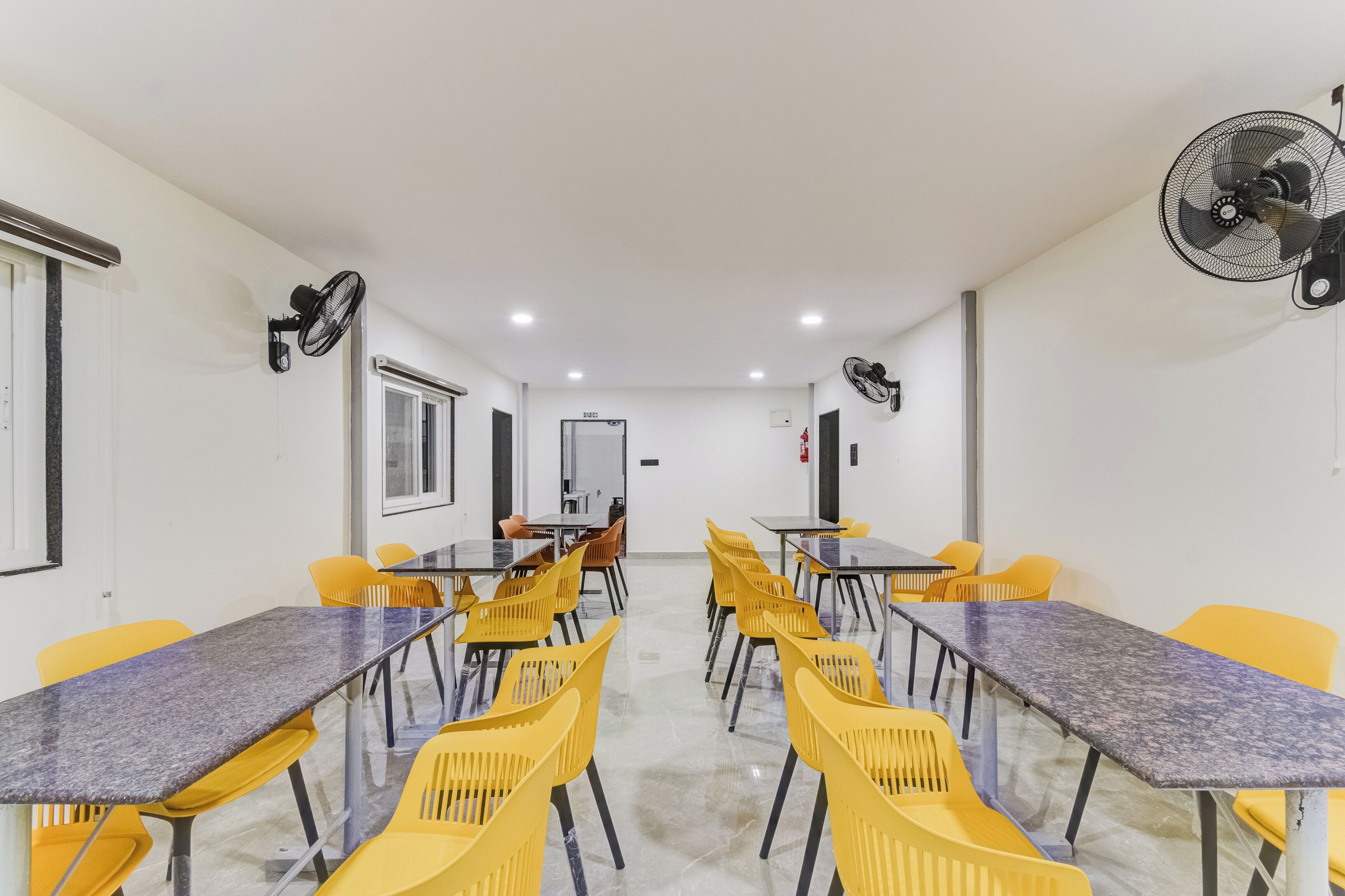 Sala de conferências com mesas, cadeiras, ventiladores de teto e uma porta ao fundo.