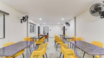 Sala de conferências com mesas, cadeiras, ventiladores de teto e uma porta ao fundo.