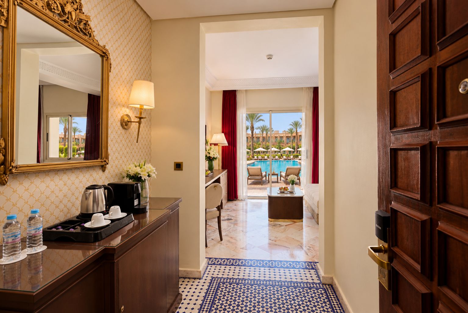Um saguão de hotel com balcão de recepção, um espelho grande, vista para área de piscina externa e piso estampado.
