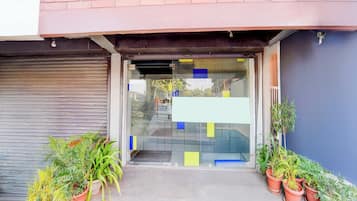 Entrada de um edifício com uma persiana fechada, porta de vidro com adesivos e plantas em vasos no chão.