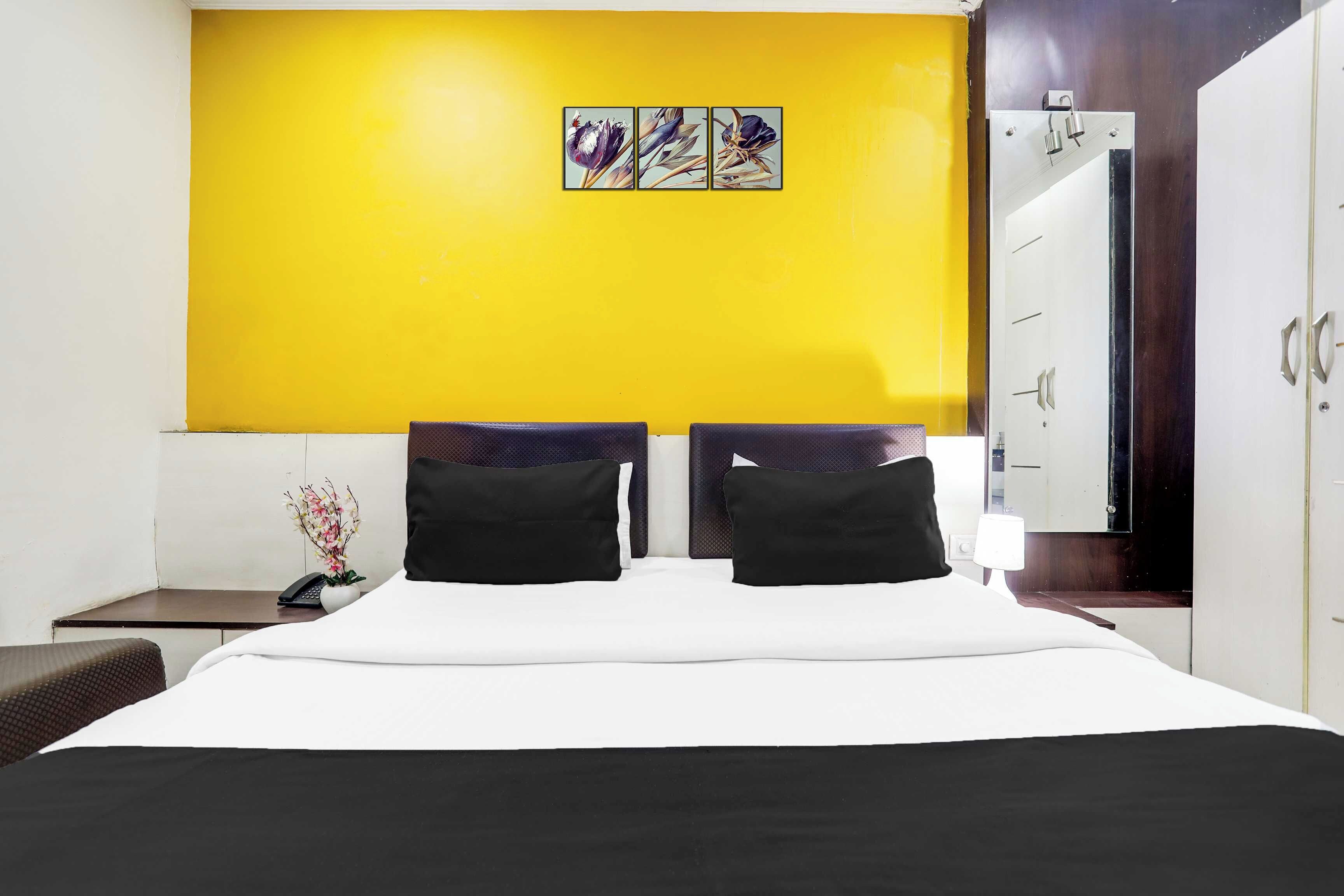 Quarto de hotel com cama, dois travesseiros pretos, mesa de cabeceira com telefone e flores, guarda-roupa e parede amarela com um quadro.