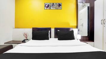 Quarto de hotel com cama, dois travesseiros pretos, mesa de cabeceira com telefone e flores, guarda-roupa e parede amarela com um quadro.
