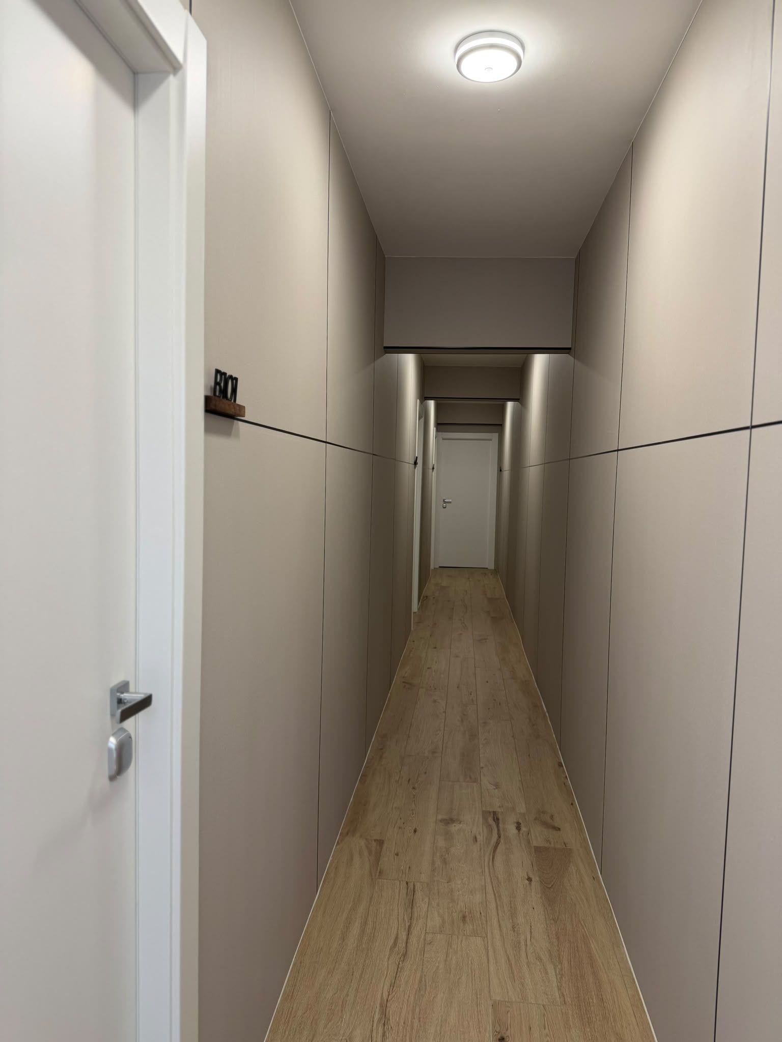 Um corredor estreito com piso de madeira, paredes brancas e uma porta à esquerda.