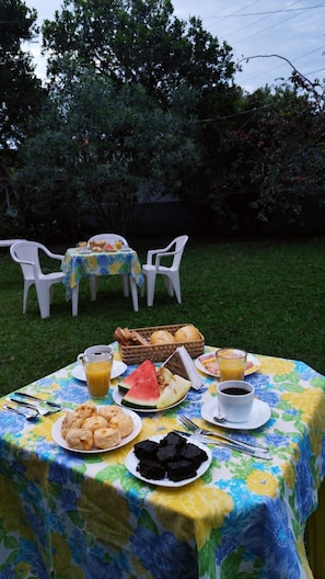 Uma área de refeições ao ar livre com mesa posta para o café da manhã, incluindo pratos de comida, bebidas e uma cesta de pão.