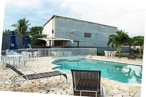 Piscina com espreguiçadeiras e mesas, cercada por um terraço de pedra e um edifício ao fundo.