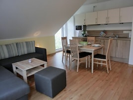 Um espaço de estar moderno com sofá, mesa de jantar, cadeiras, cozinha compacta e janela que permite entrada de luz natural.