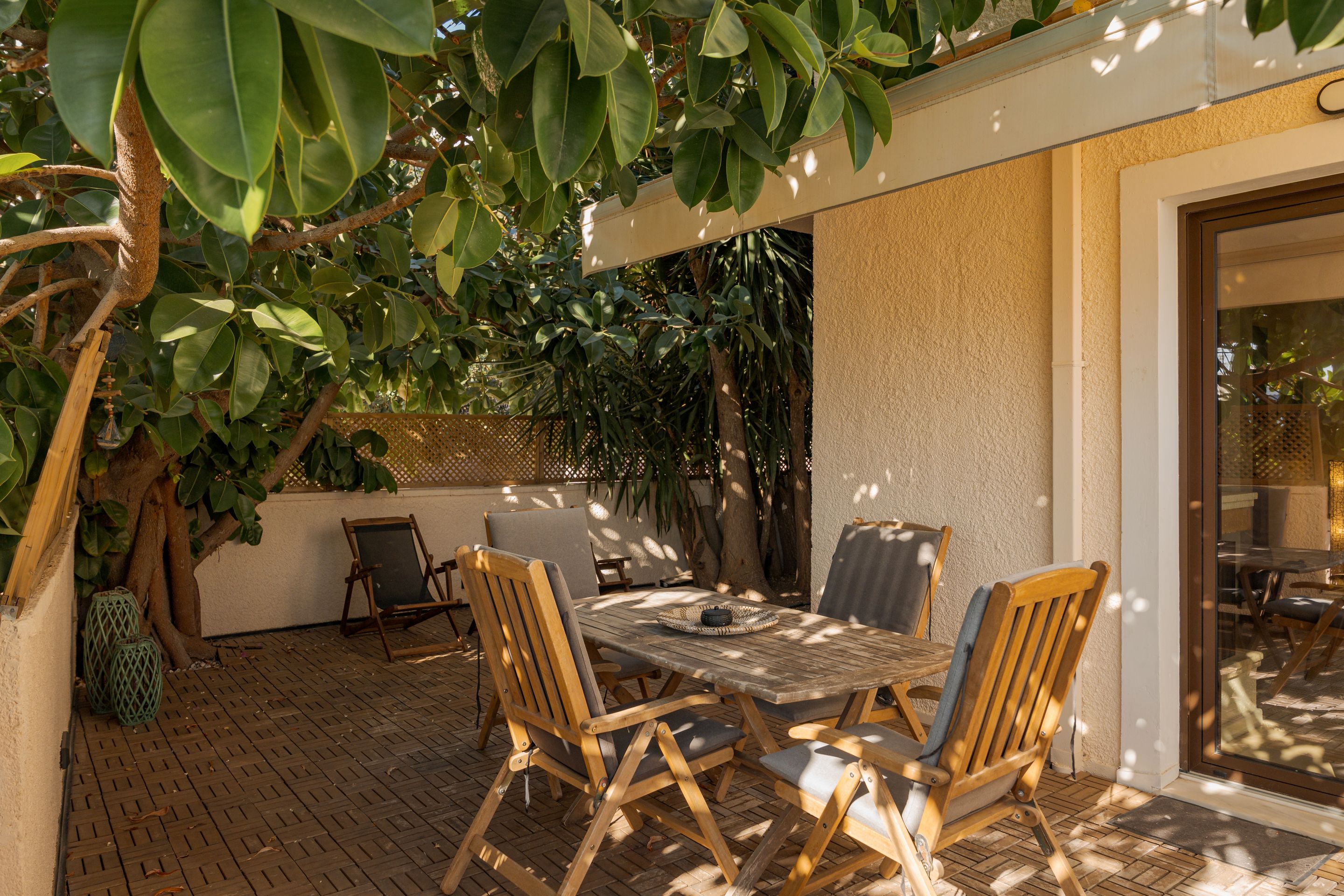 Um pátio externo com mesa e cadeiras, cercado por vegetação e uma parede branca.