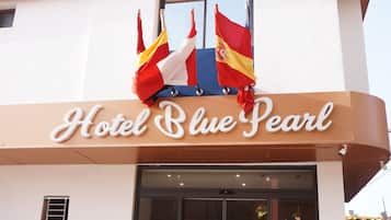 Entrada de um hotel com uma placa indicando 'Hotel Blue Pearl' e duas bandeiras sobre a entrada.