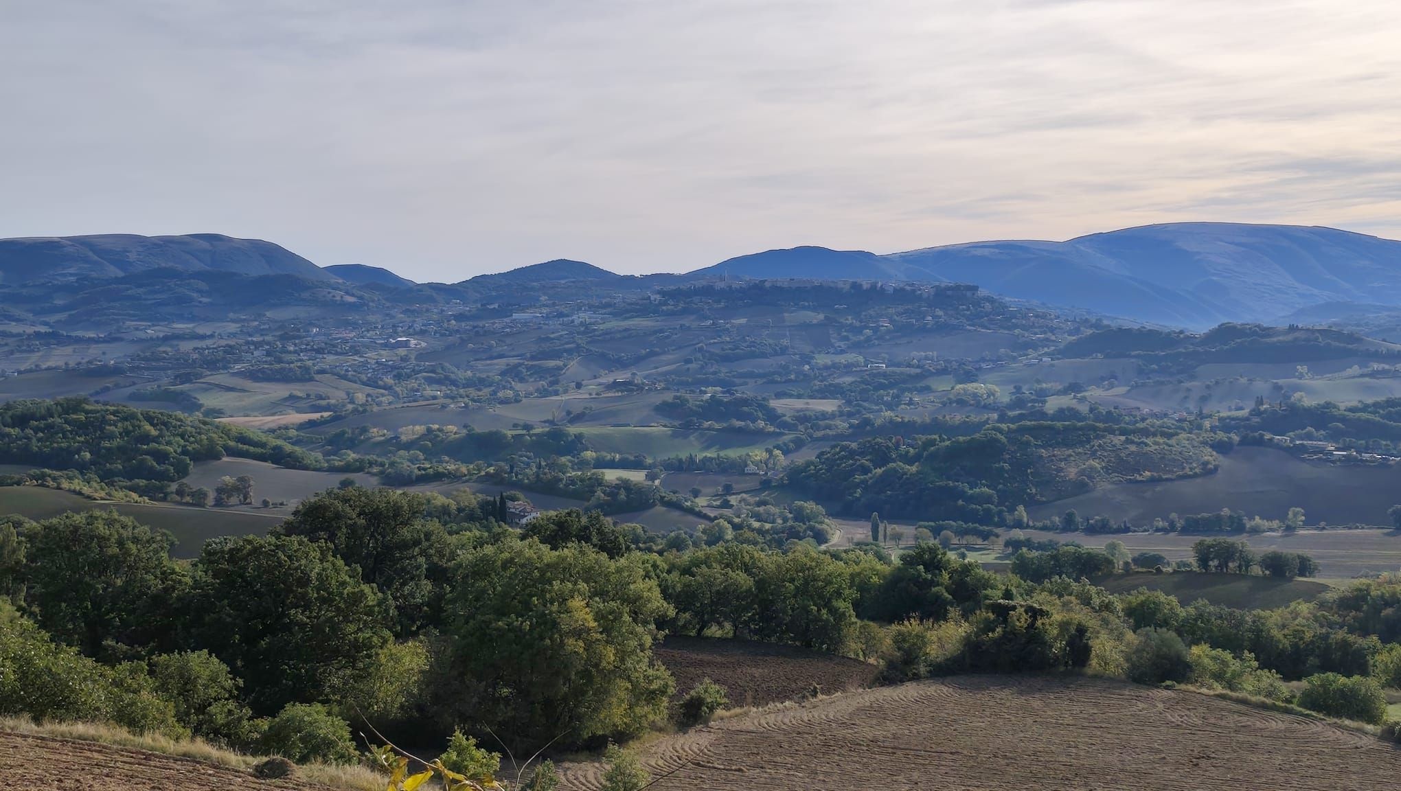 Vista panorâmica de uma paisagem rural com colinas ondulantes, árvores dispersas e uma pequena aldeia.
