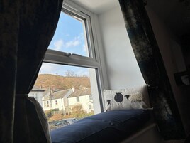 Quarto com janela vista para área residencial, cama com colchão azul e travesseiro com desenho de ovelha.