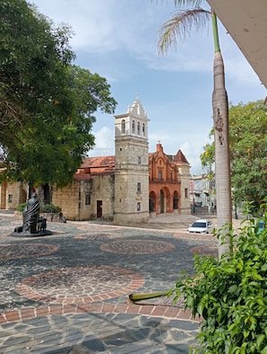 Uma praça com um edifício histórico, com uma torre de sinos e arcos, cercada por árvores e uma estátua.