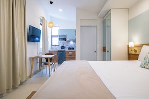 Quarto de hotel com cama, escrivaninha, cadeira, televisão, cozinha e porta.
